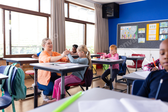 Female teacher assisting young girl with schoolwork in classroom, other ...