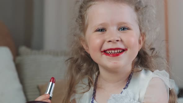 Cheerful little girl cyild paints her lips with bright red lipstick ...