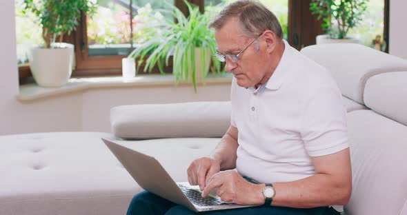 A Senior Retired Man Types on a Laptop Computer at Home on a Couch ...