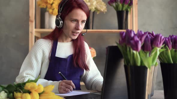 Florist in Headphones with a Microphone and an Apron Sits at a Table ...