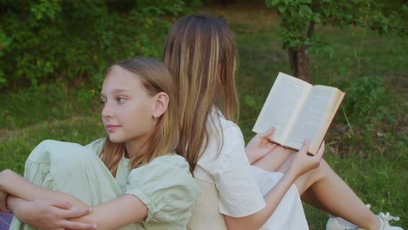 Relaxed Teenager Girls Sitting Back to Back Reading Book on Green Grass ...