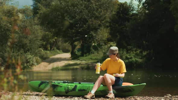 A young woman approaches the kayak and sits down, pouring a drink from ...