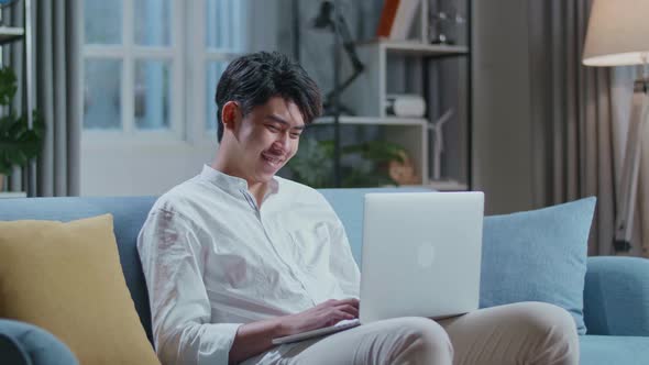 Smiling Asian Man Typing On Laptop Computer While Sitting On Sofa In ...